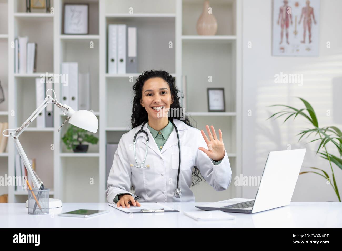 Friendly Hispanic female doctor sitting in her office, waving at the ...