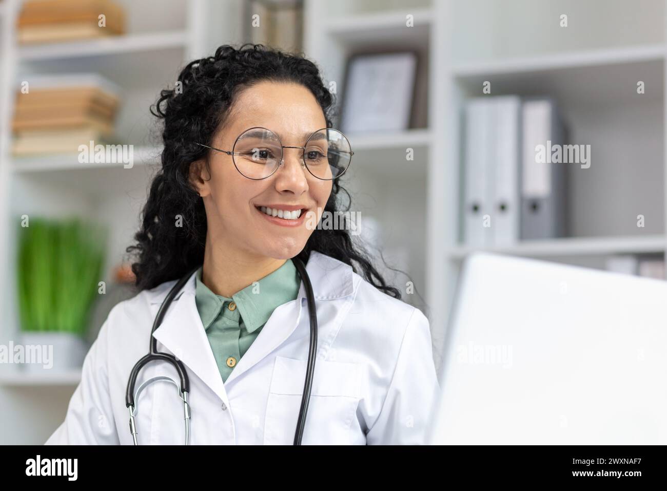 A joyful Latina doctor wearing glasses smiles cheerfully at work ...