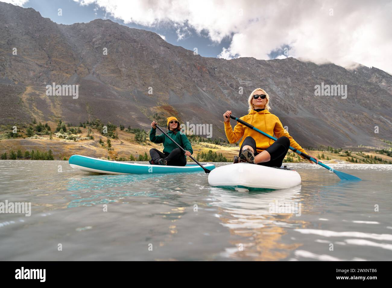 Two surfers girls sits and walks on sup boards at calm mountain lake ...