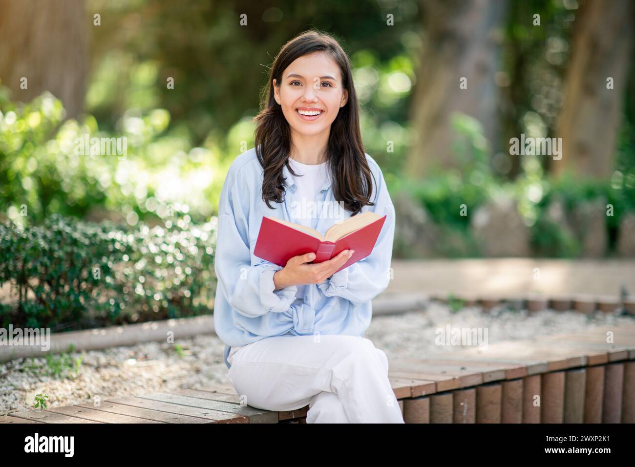 Enthusiastic young reader sits on a park bench with a beaming smile ...