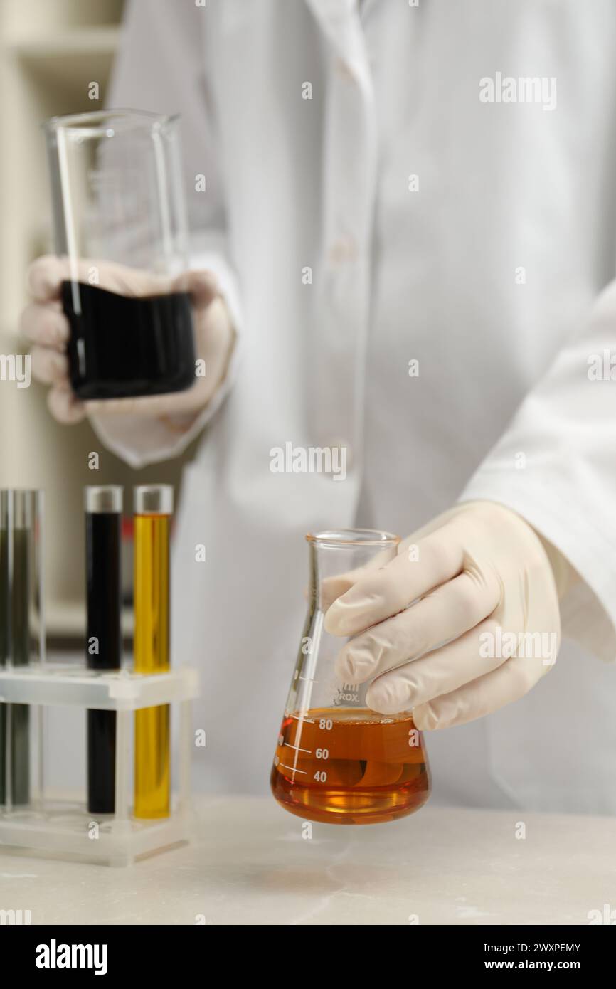 Laboratory Worker Holding Beaker And Flask With Different Types Of Crude Oil At Light Marble 