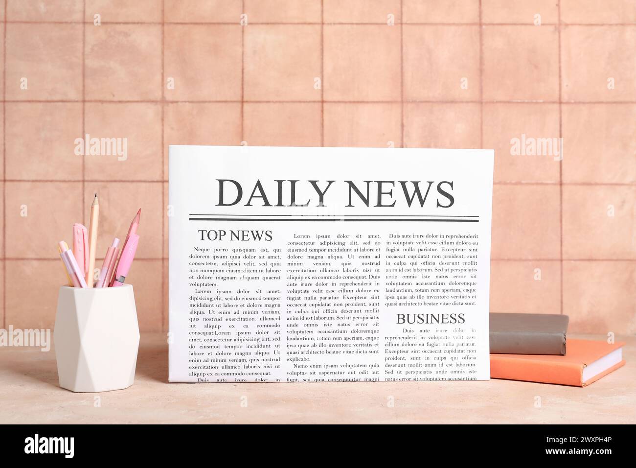 Newspaper with books and cup of pens on table against beige tile ...