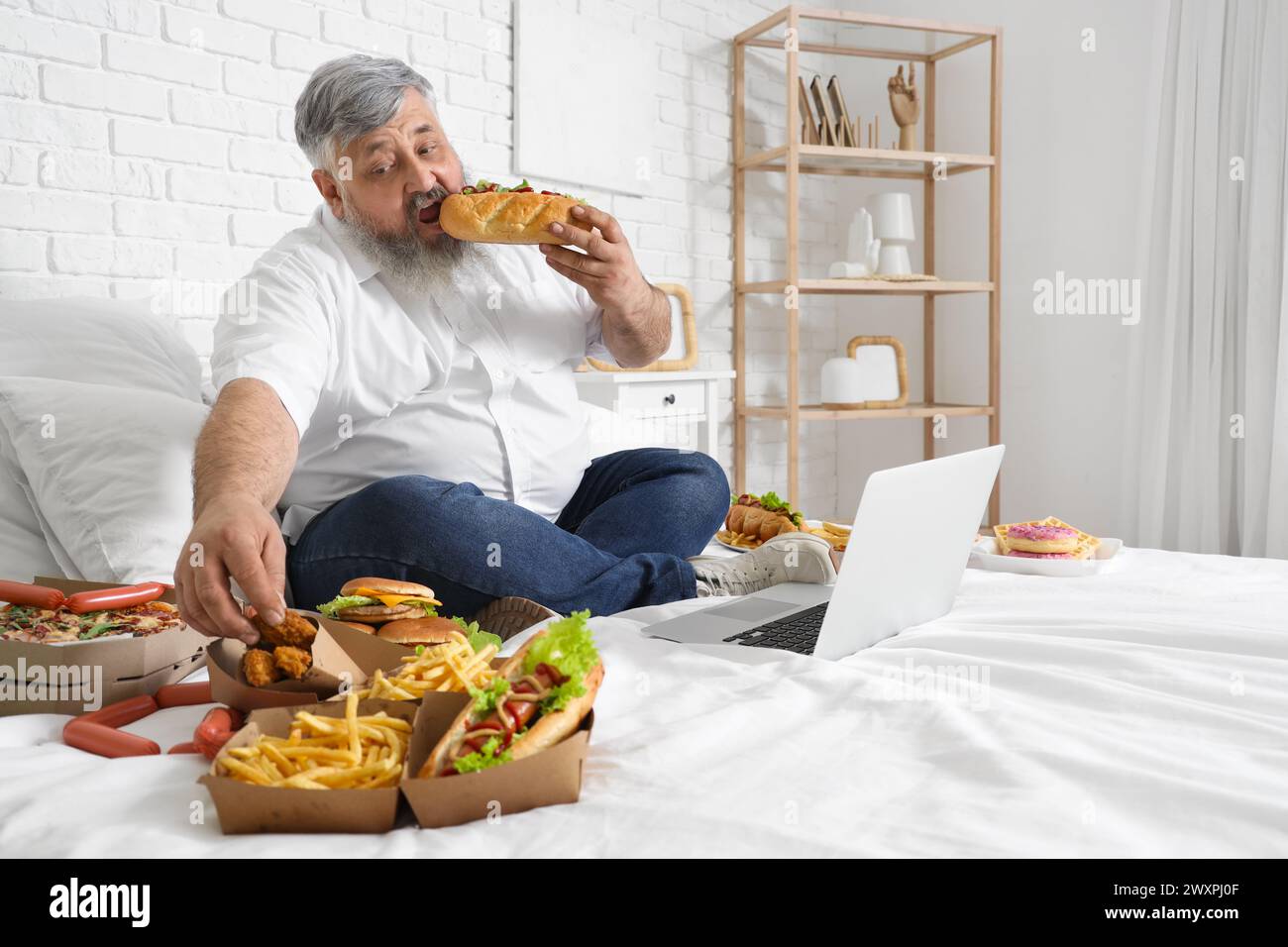 Overweight mature man with unhealthy food and laptop sitting in bedroom ...