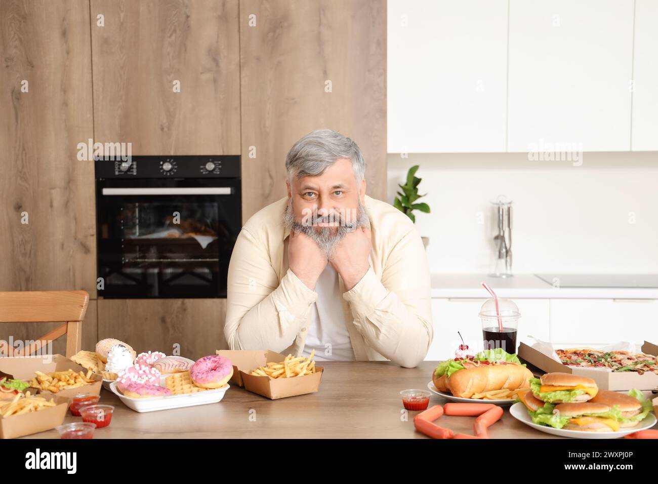 Overweight mature man at table full of unhealthy food in kitchen ...