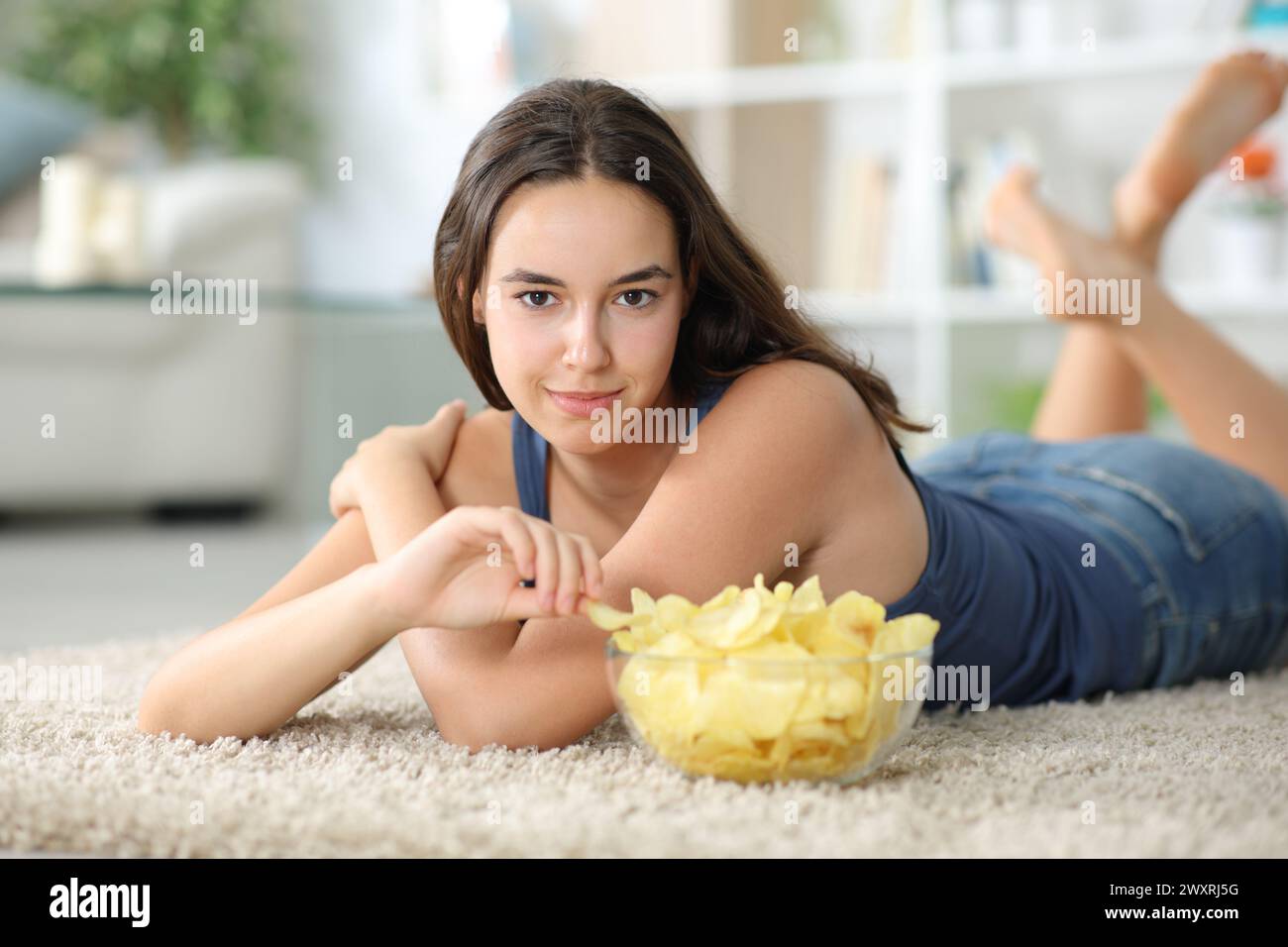 Beautiful woman looks at you eating potato chips lying on a carpet at ...