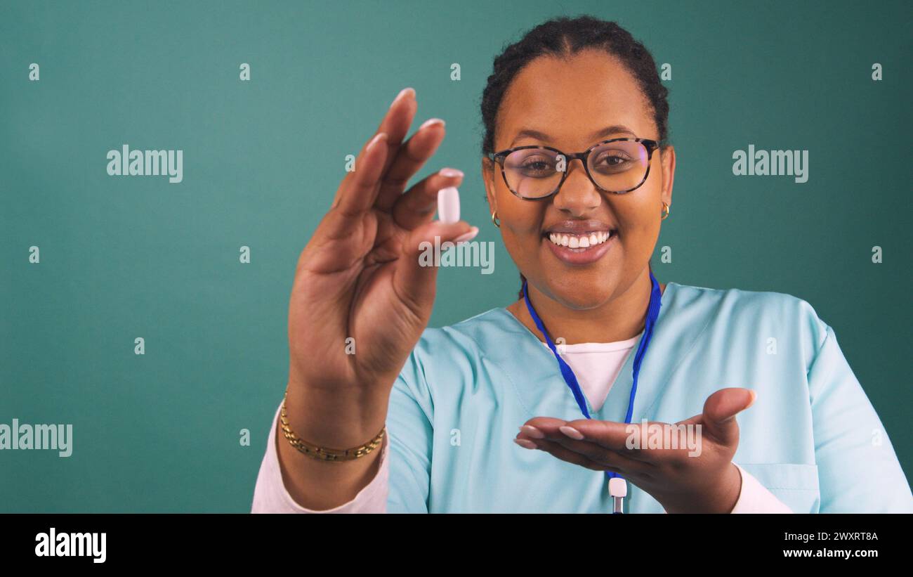 Young Black female nurse talking and holding pill, smiling in blue ...