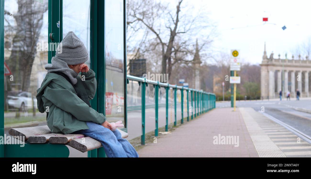 crying lonely child, wiping eyes with hand, in warm cloth at bus stop ...