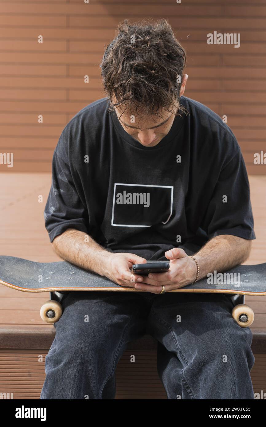 A skateboarder takes a break to check his smartphone, sitting on his ...