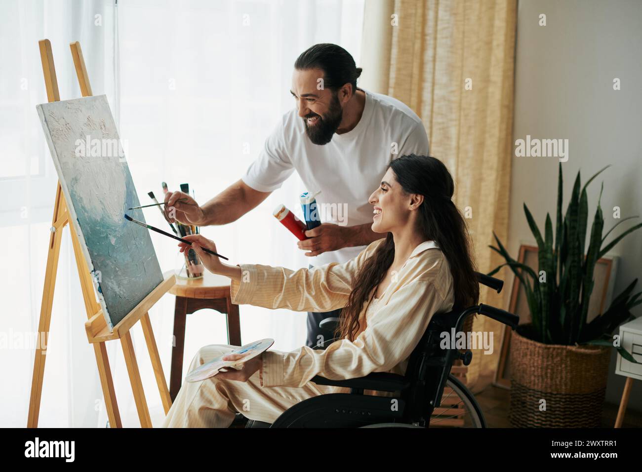 good looking bearded man watching disabled beautiful wife on wheelchair painting on easel at ...