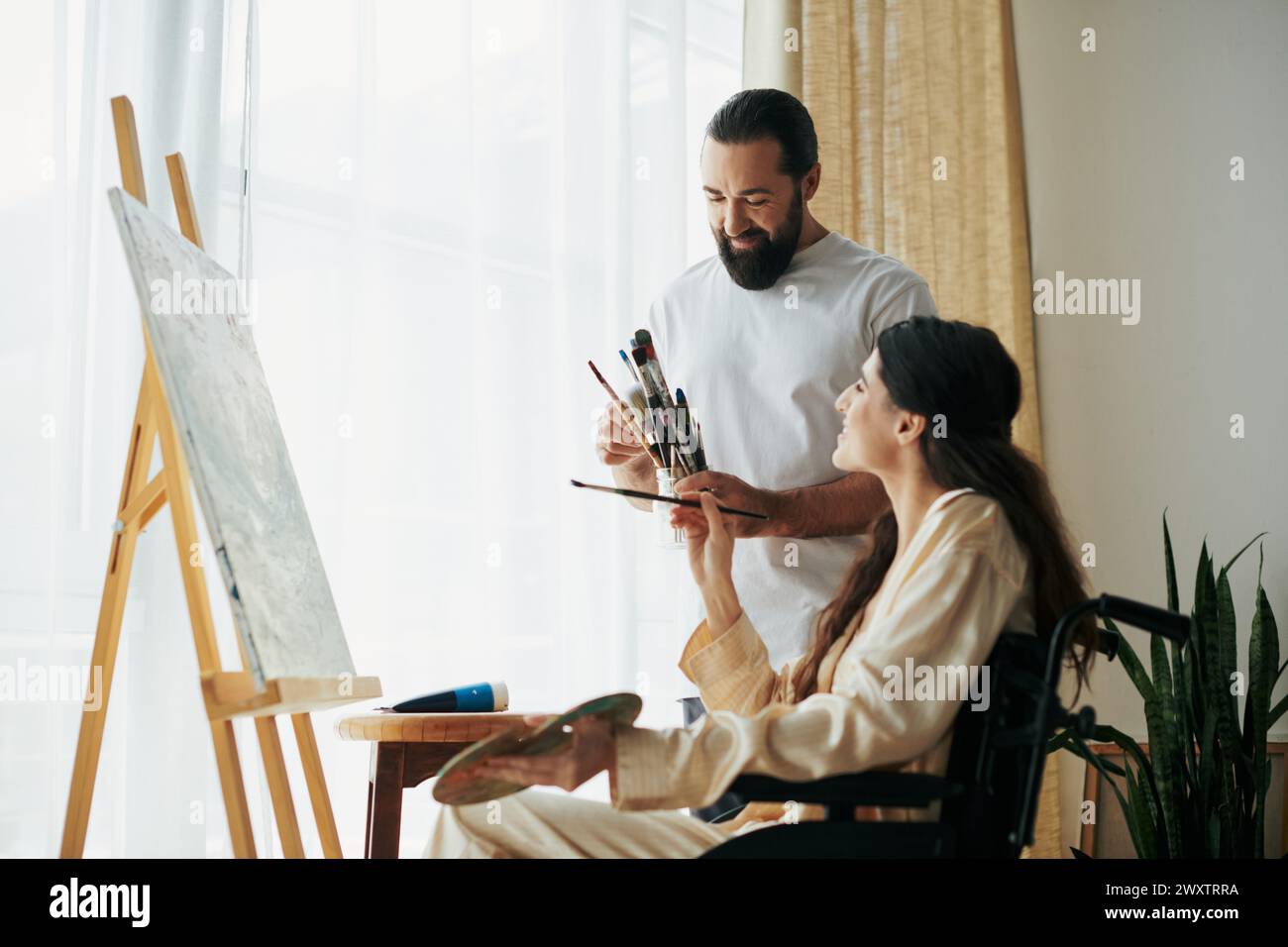 good looking bearded man watching disabled beautiful wife on wheelchair painting on easel at ...