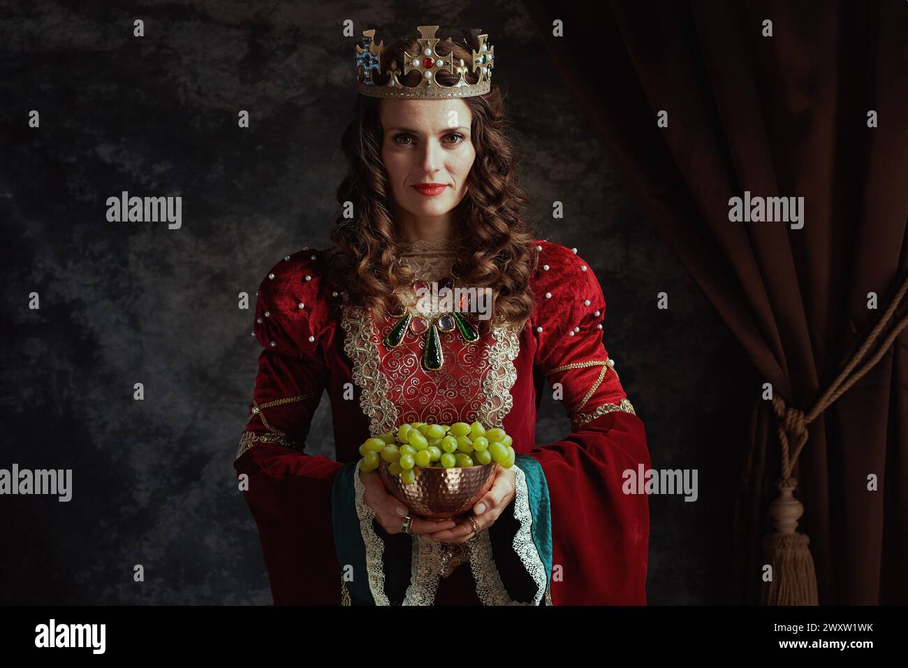 smiling medieval queen in red dress with plate of grapes and crown on ...