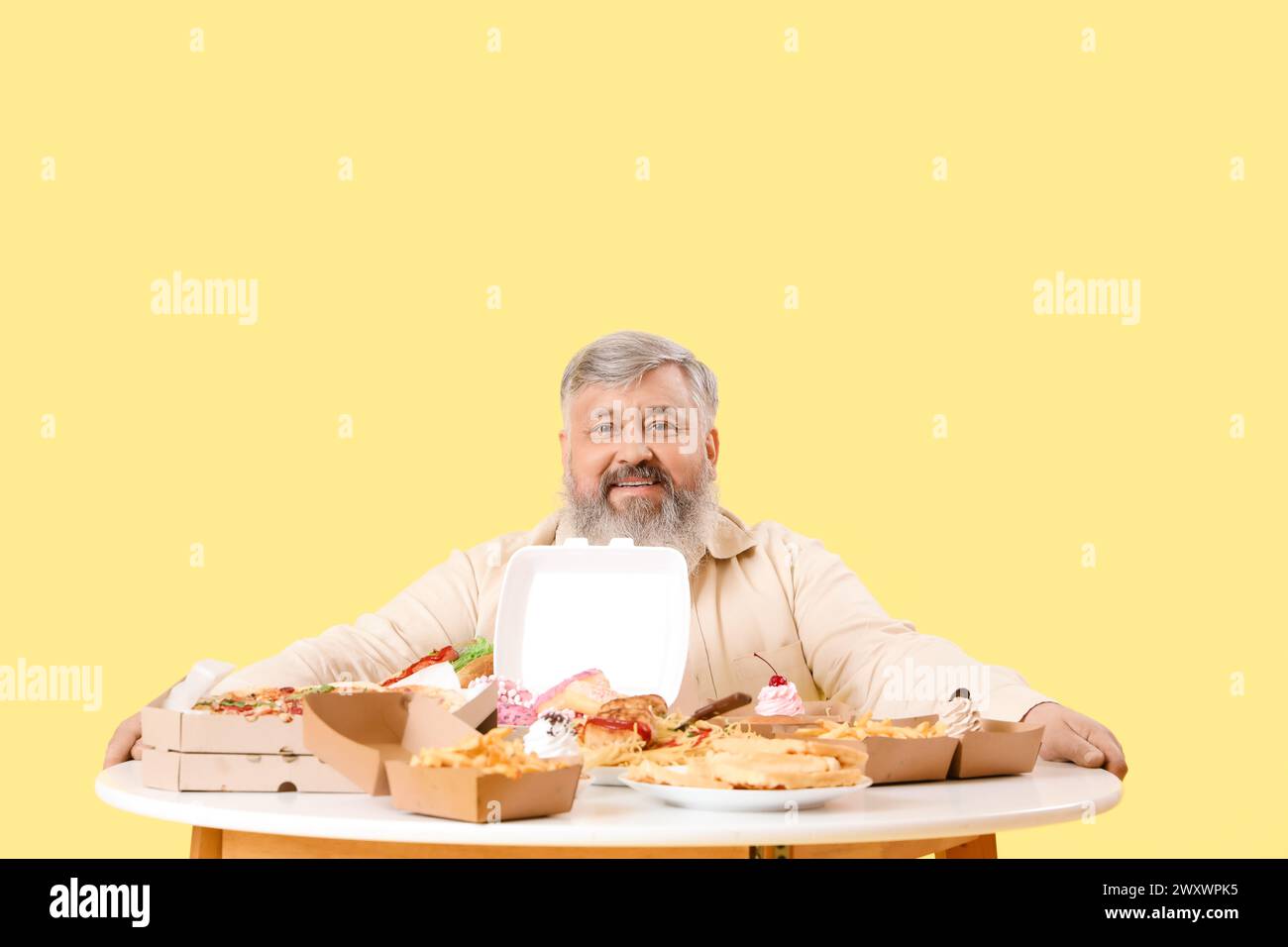 Overweight mature man with unhealthy food on table against yellow ...