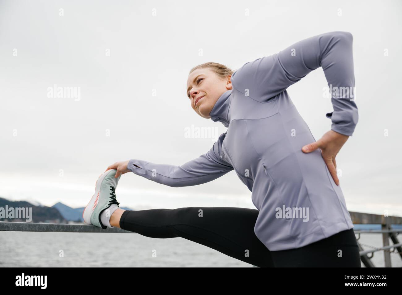 A young athletic woman performs stretching exercises on a cloudy day, with a tranquil water ...