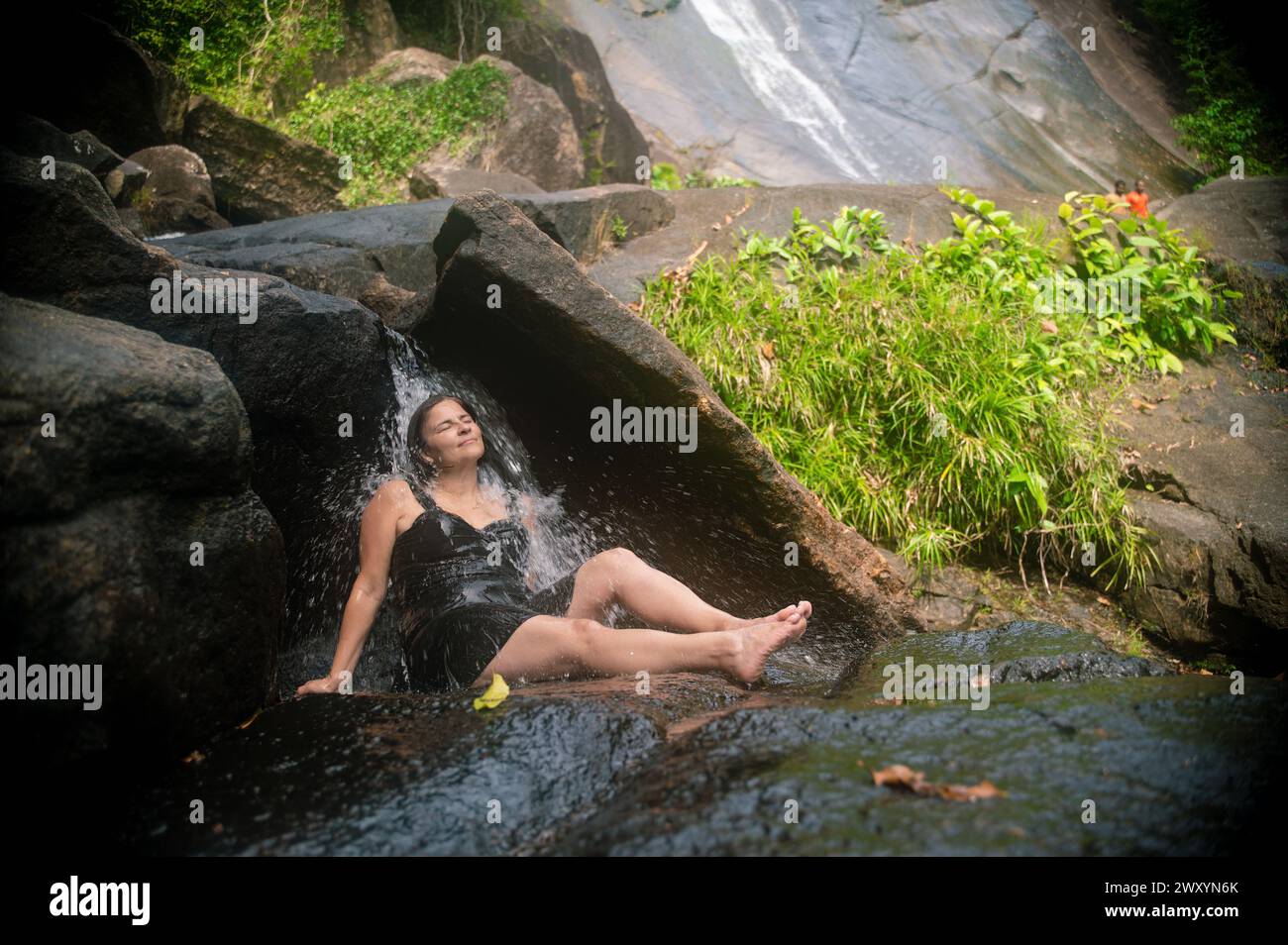 A woman reclines on a rock, feeling the gentle flow of a waterfall in ...