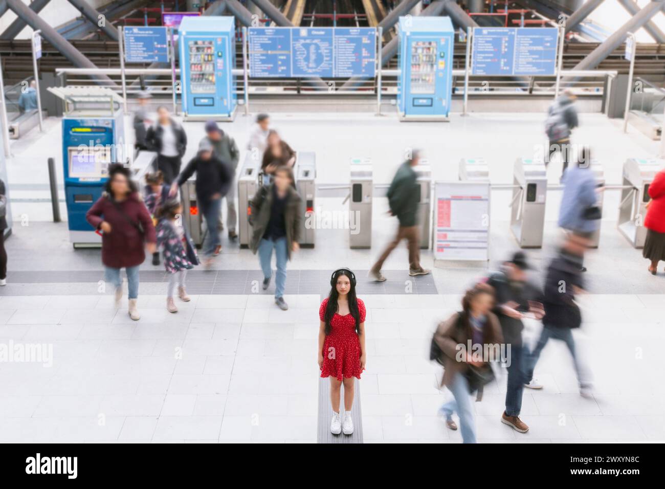 A young woman in a red dress remains motionless as blurred figures rush ...