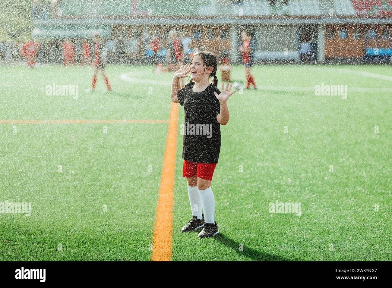 A young girl in soccer attire enjoys the cooling rain on a lush green ...