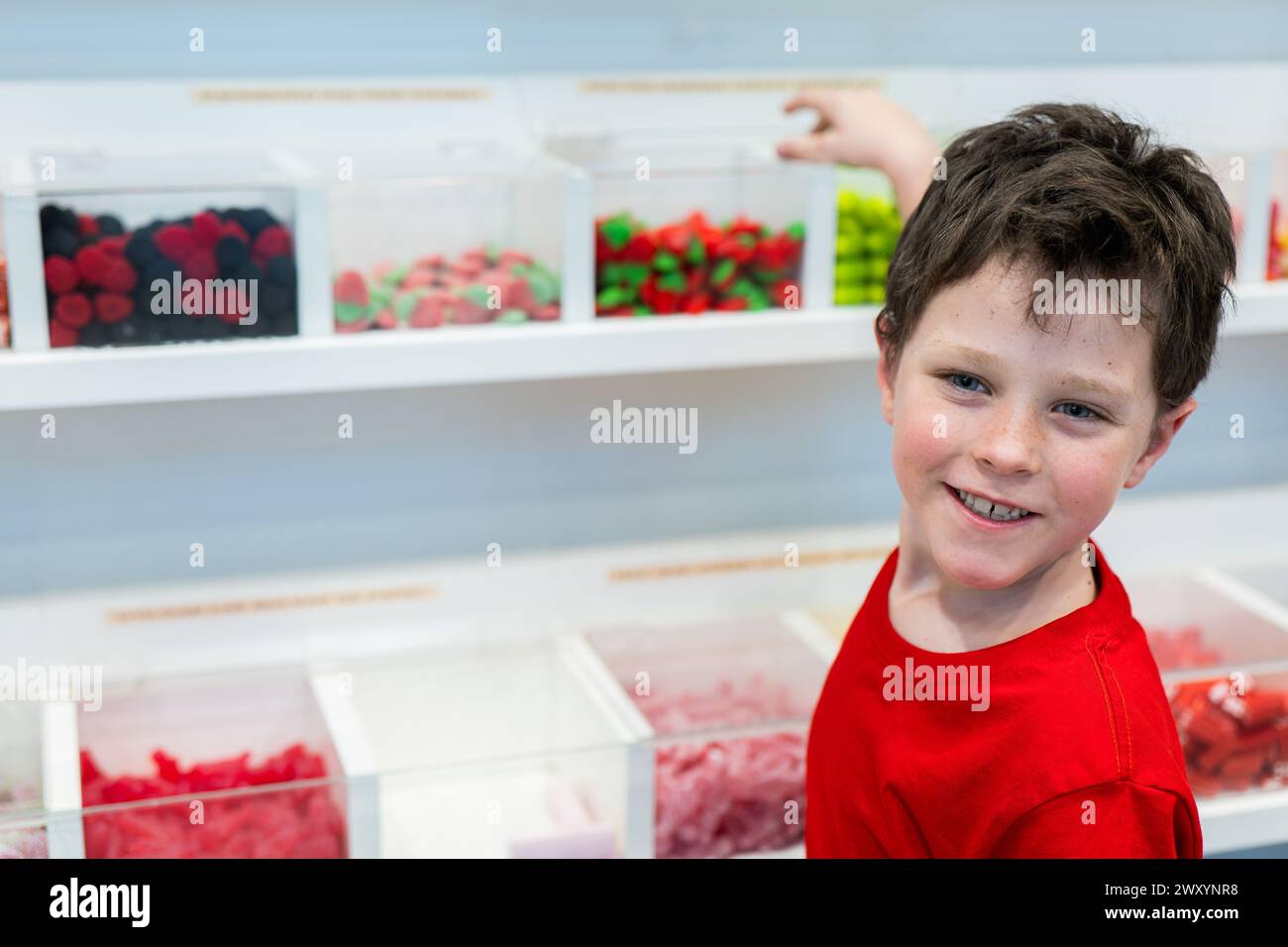 A young boy in a red shirt is smiling in front of a candy display ...