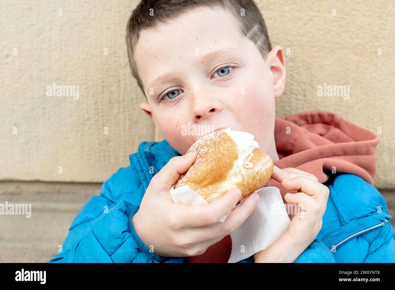 A young boy with blue eyes eats a cream-filled pastry, with traces of ...