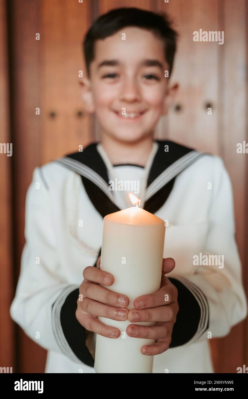 A content young boy in First Communion dress holds a candle, its flame ...