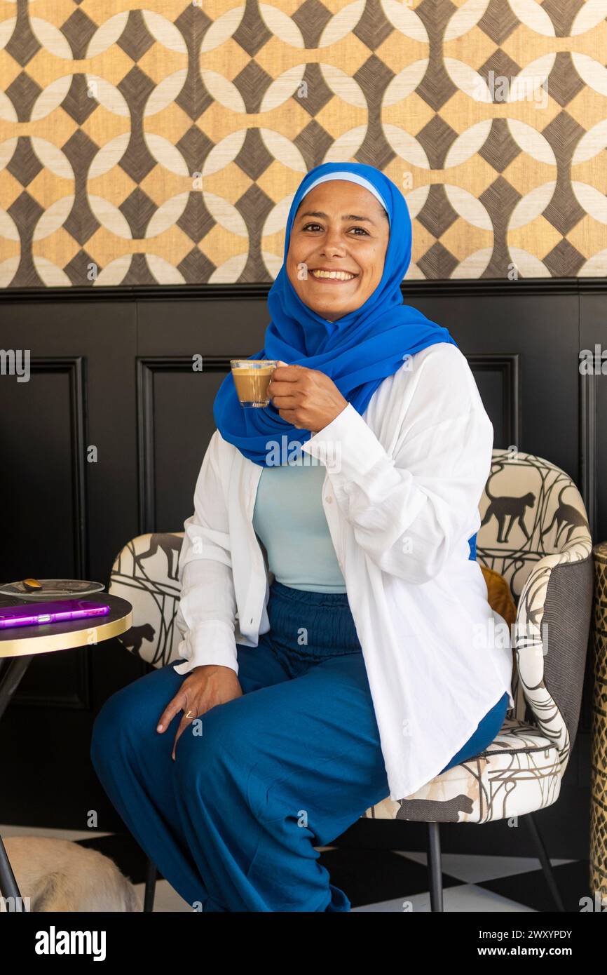 Cheerful multiethnic woman wearing a blue hijab sips coffee in an ...