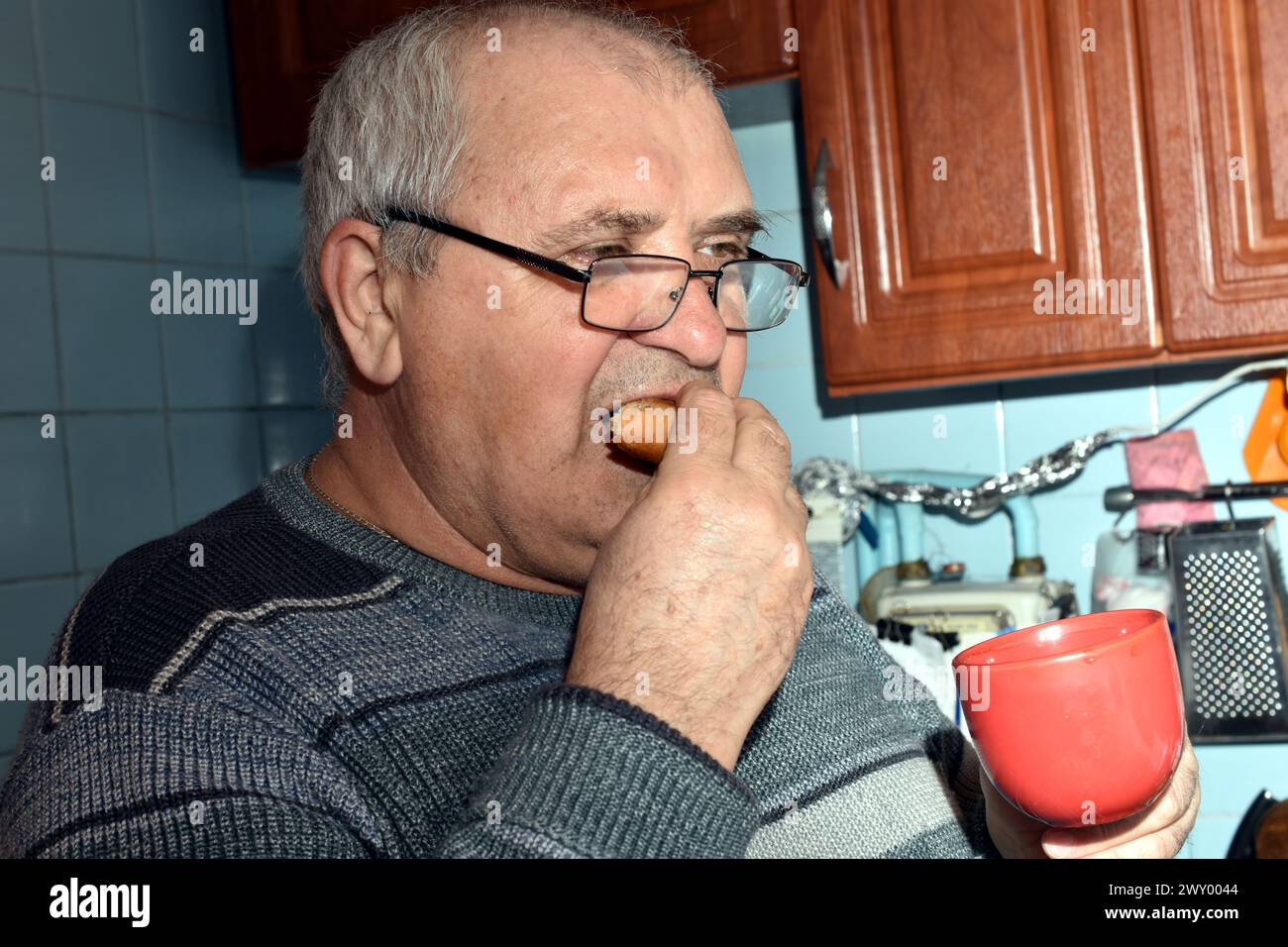 The picture shows an elderly man, he is eating a bun and holding a cup ...