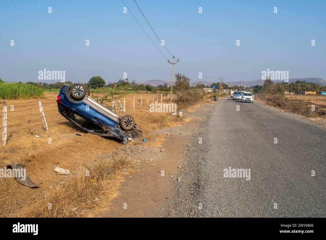 A blue car overturned beside a rural road with distant traffic and ...