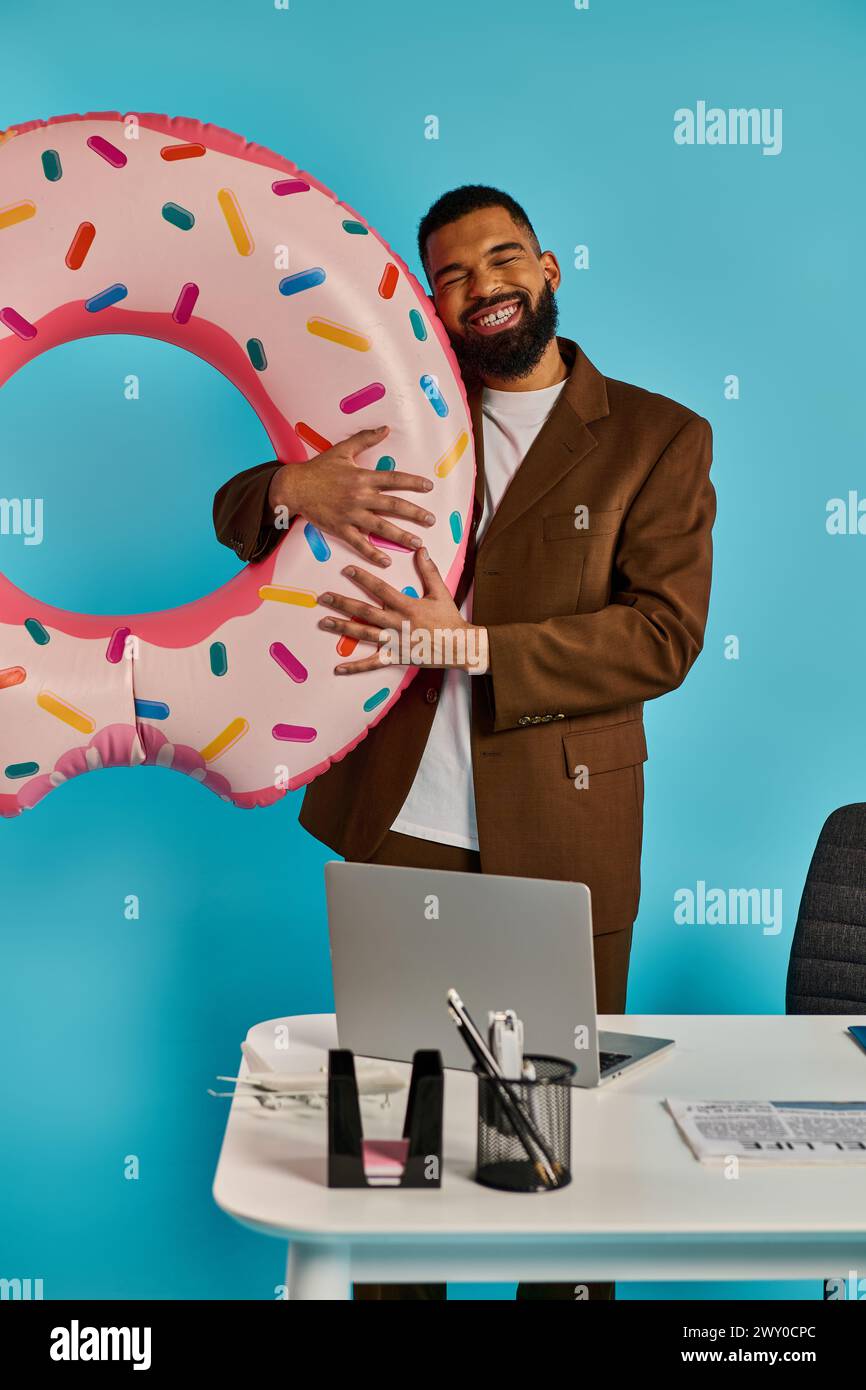 A man is holding a massive donut in front of a laptop, seemingly ...