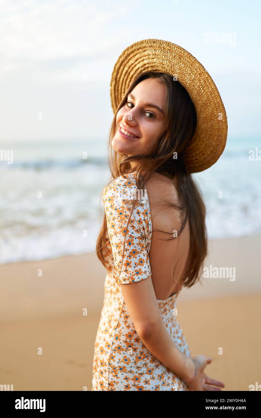Smiling young woman in floral summer dress and straw hat strolls along ...