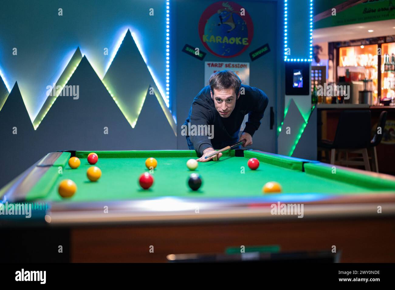 A young man dressed in black and smiling playing pool in a dark ...