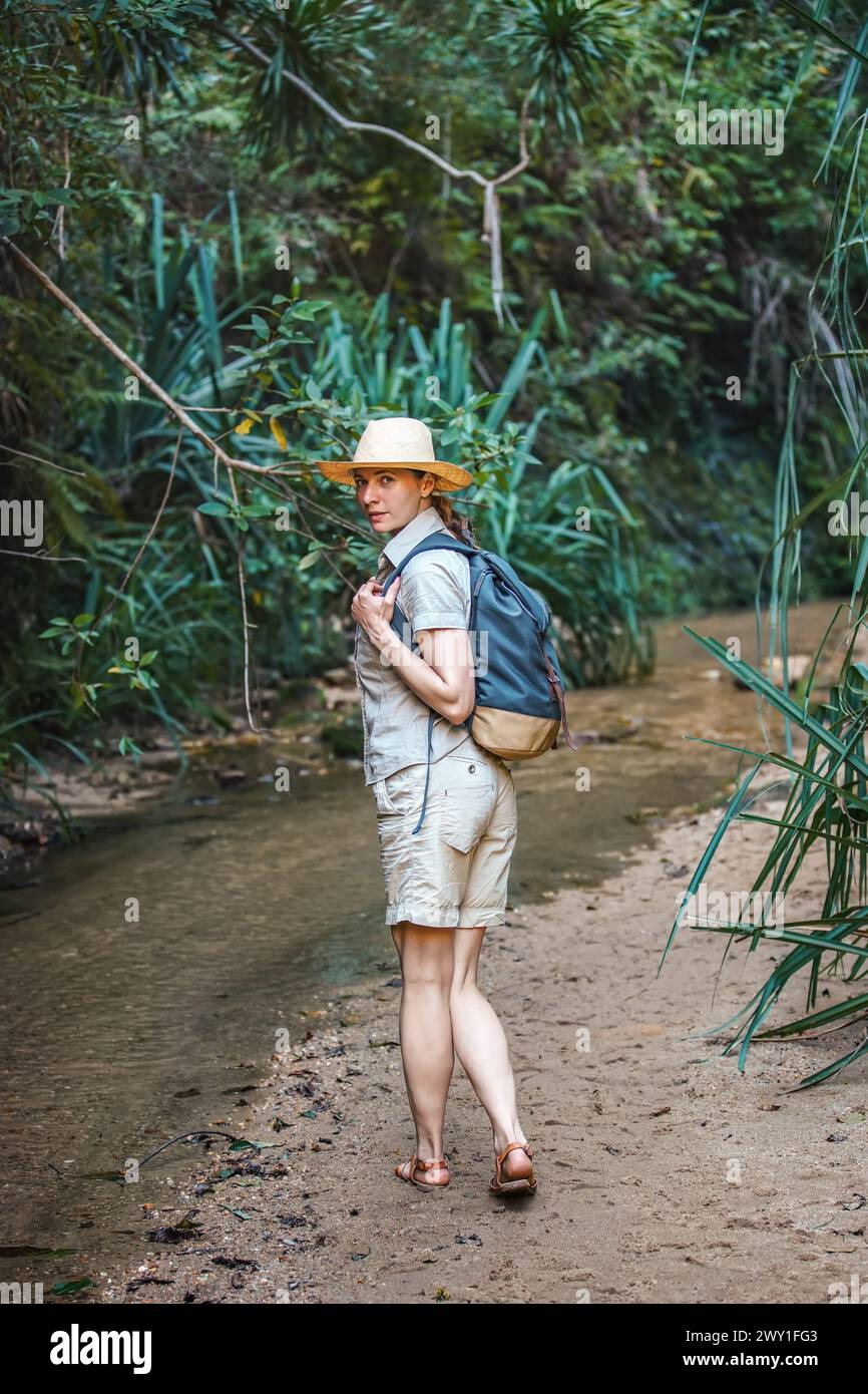 Young woman in beige shirt, shorts and straw haw, backpack on shoulders ...