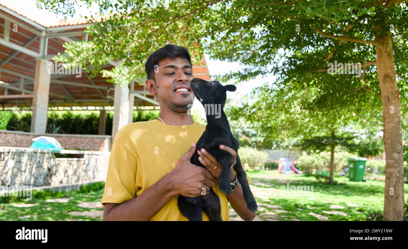 A gentle Indian man with a warm smile cradles a tiny black baby goat in ...
