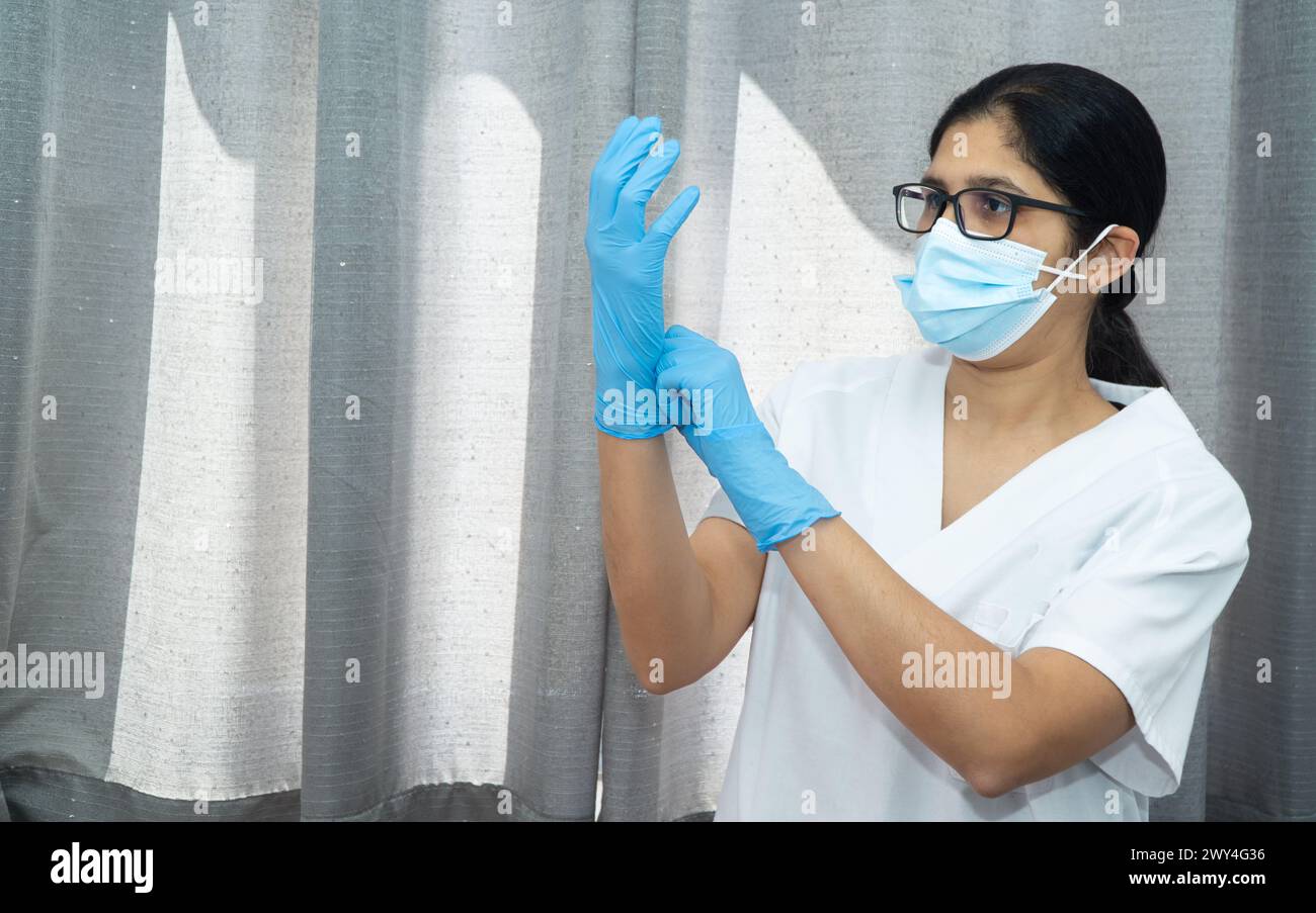 A woman wearing a white lab coat and blue gloves is putting on a mask ...