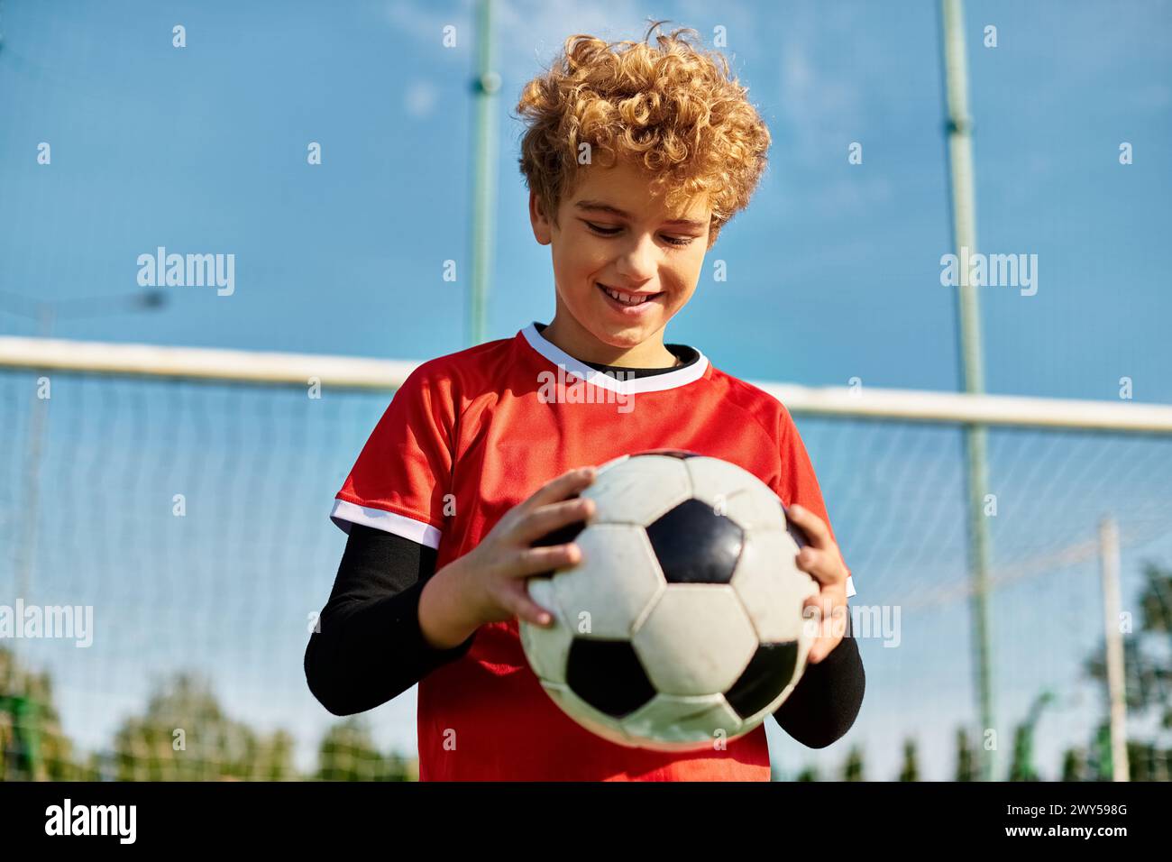 A young boy stands in front of a goal, holding a soccer ball. He ...