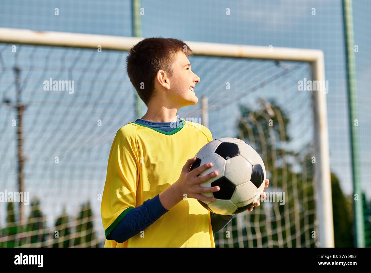 A young boy stands in front of a soccer goal, holding a soccer ball. He ...