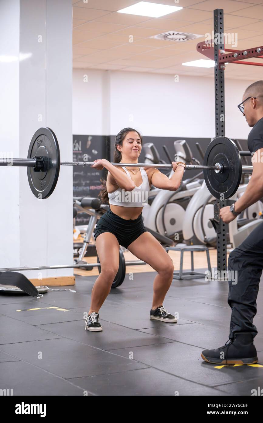 Fitness enthusiast performs a barbell squat in a gym under the watchful ...