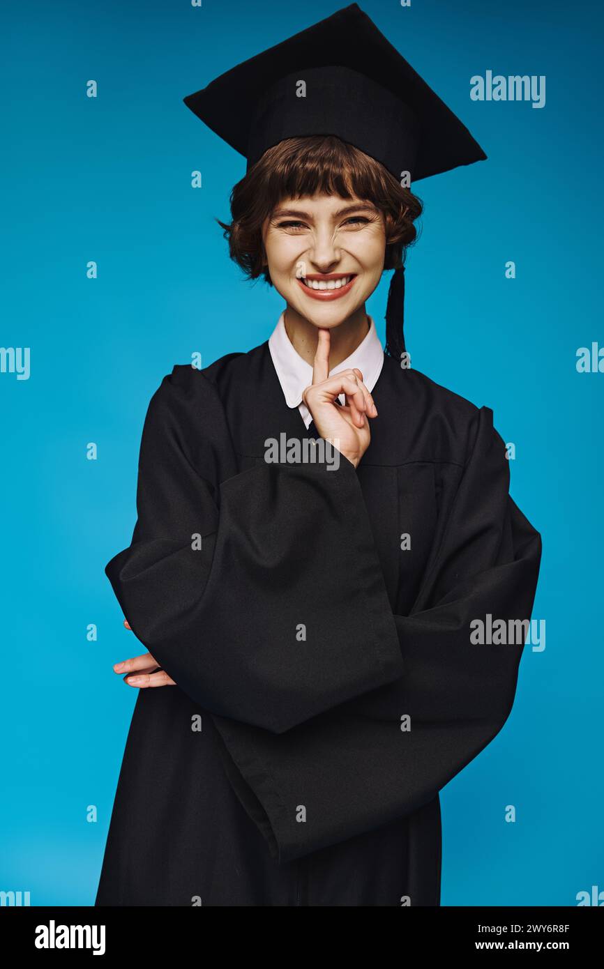 positive graduated college girl in gown and cap smiling on blue ...