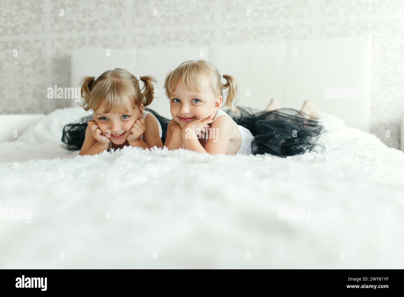 Two young female children are reclining on a white bed together in a ...