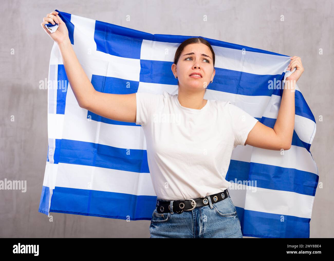 Sad young woman holding Greece flag against unicoloured background ...