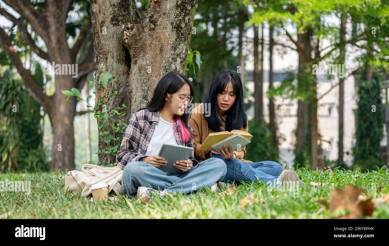 Two young Asian female students sit on the grass under a tree, talking and studying together in ...