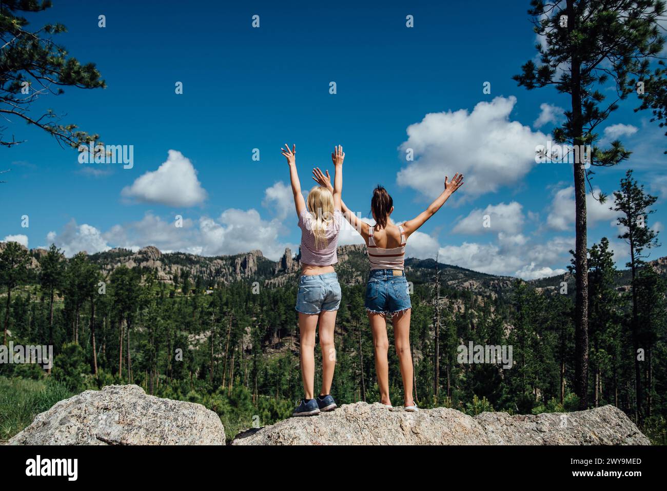 Rear view of two teen girls with arms to sky overlooking mountains ...