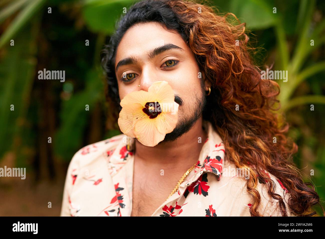 Dark-skinned man with curly long hair poses in nature, flower in mouth ...