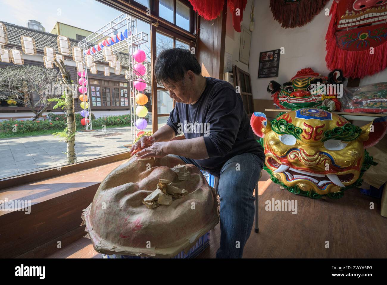 An artisan is hand making a Chinese mask in a sunlit workshop ...