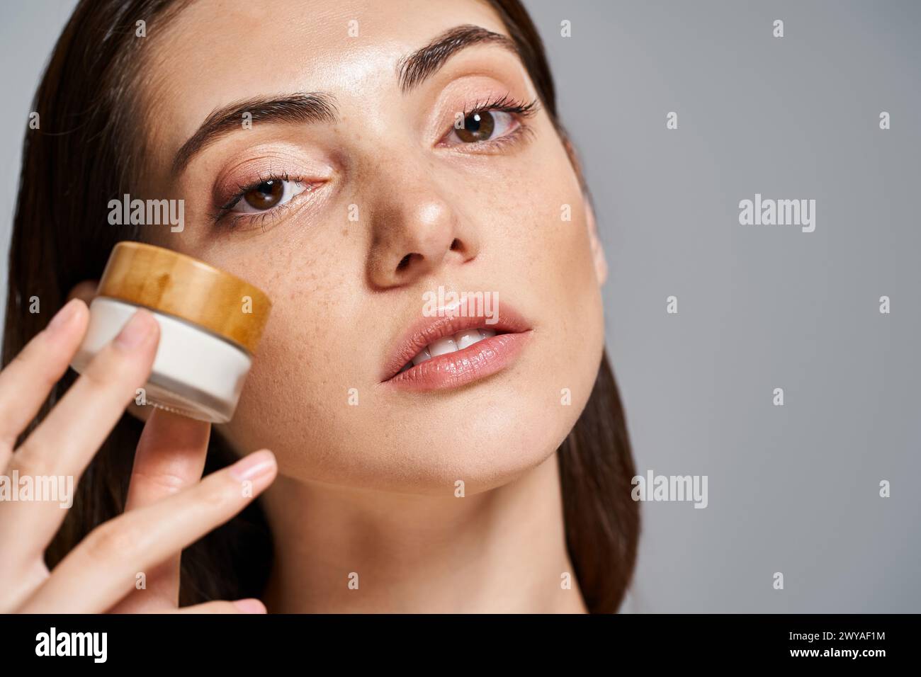 A young Caucasian woman with brunette hair applying cream from a jar to ...