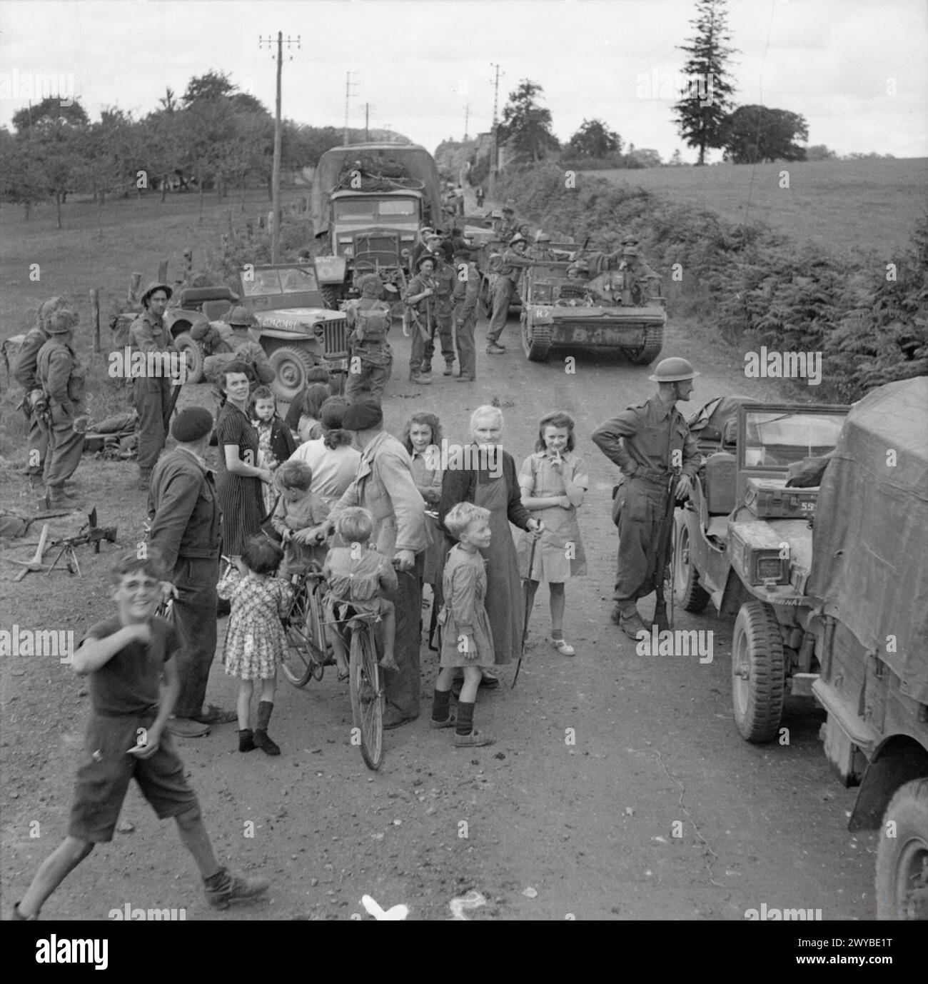 Local villagers greet a British column of trucks, jeeps, and carriers ...