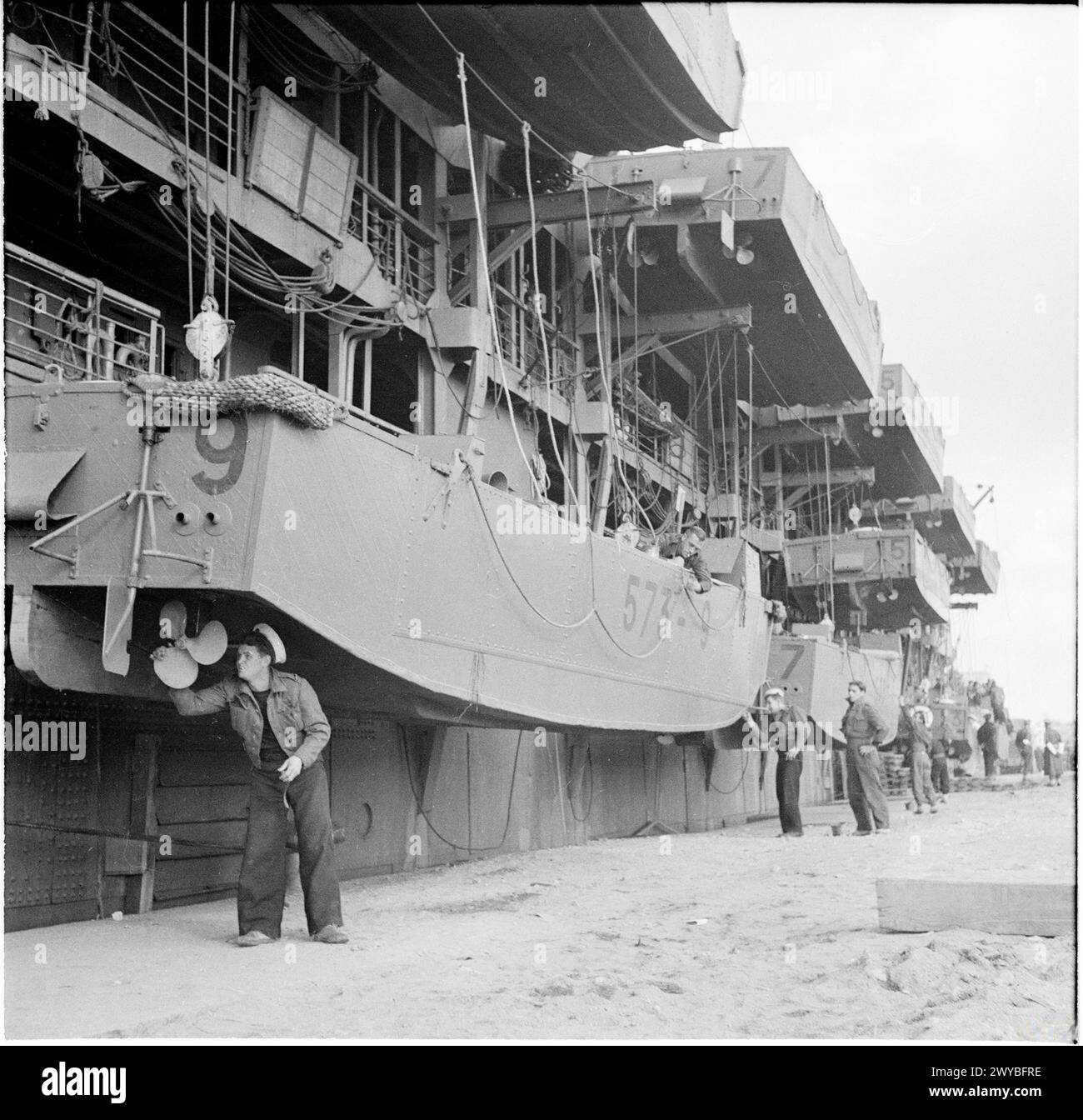 Naval personnel inspect landing craft suspended from derricks on ...