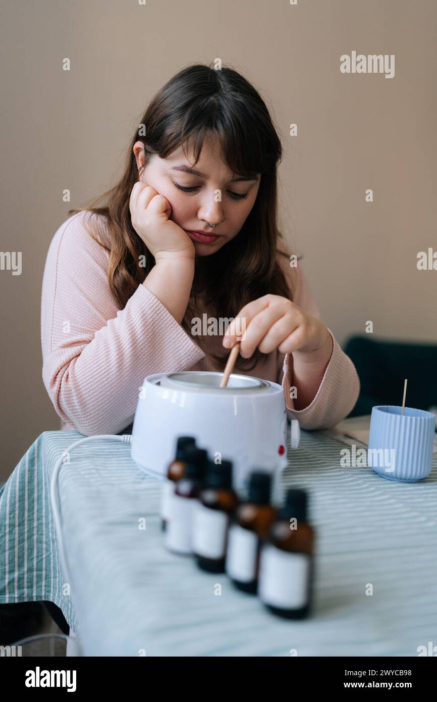 Vertical portrait of bored female artisan craftperson stirring melted ...