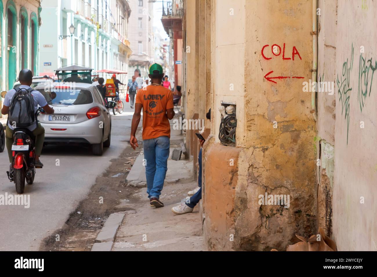 Cuban people and traffic on a city street, a word in the wall reads COLA (line-up, waiting in ...