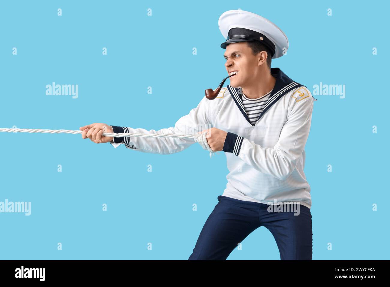 Angry young sailor with smoking pipe pulling rope on blue background ...