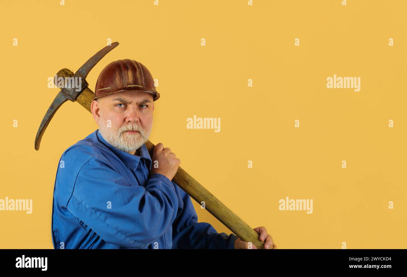 Serious bearded male builder in coveralls and hard hat with pick axe ...