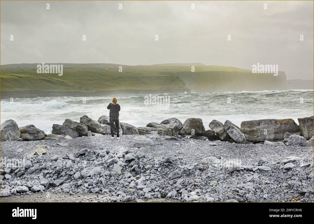 Female stood on a Stoney beach looking out to sea at some cliffs while ...